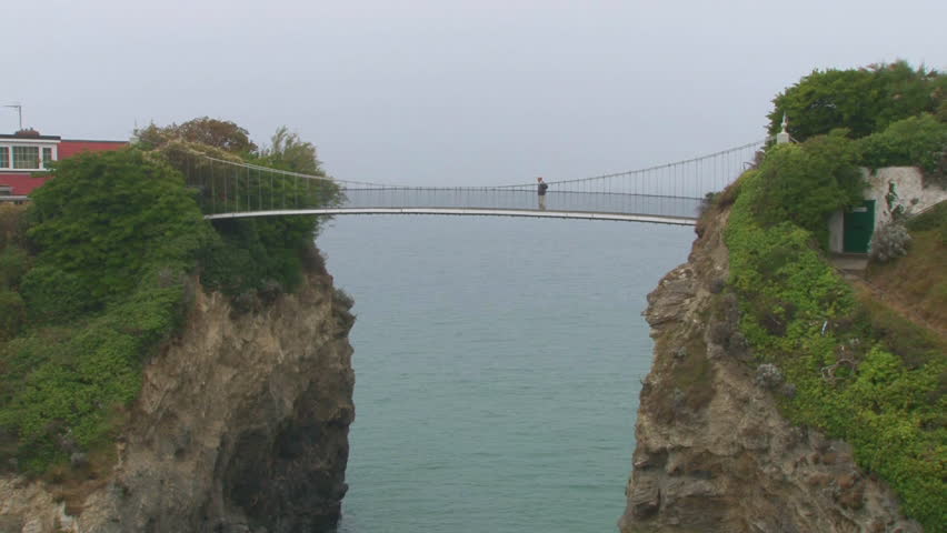 Man Walks Across A Suspended Footbridge Between Two Cliffs Over The ...